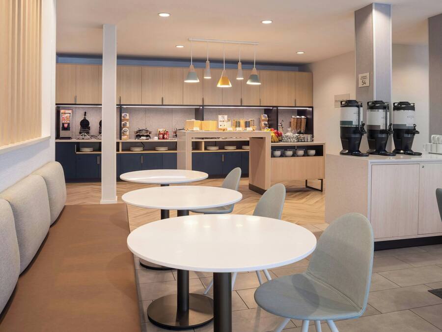 Um café moderno com mesas e cadeiras arredondadas, uma área de cozinha com máquinas de café e um espaço de estar com um sofá.