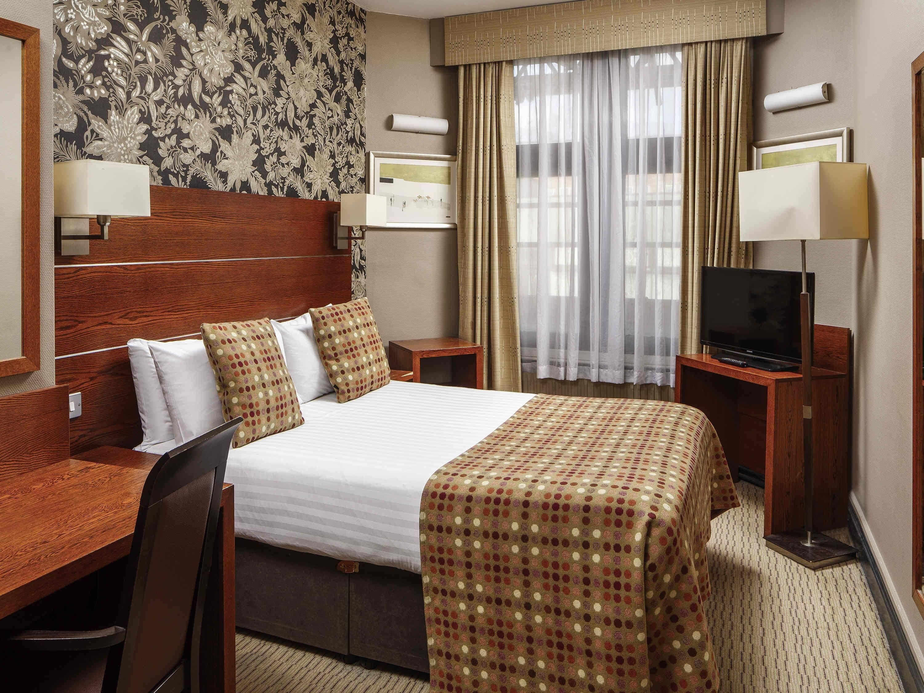 Quarto de hotel com uma cama grande, uma escrivaninha, televisão e uma janela com cortinas.