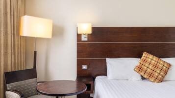Quarto de hotel com cama, uma cadeira, uma mesinha, mesa de cabeceira e duas luminárias fixadas na parede.