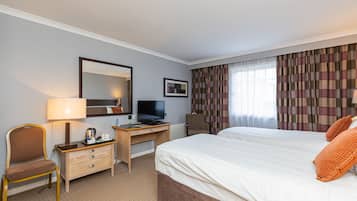 Quarto de hotel com cama, escrivaninha com televisão, cadeira, cómoda e uma janela grande com cortinas.