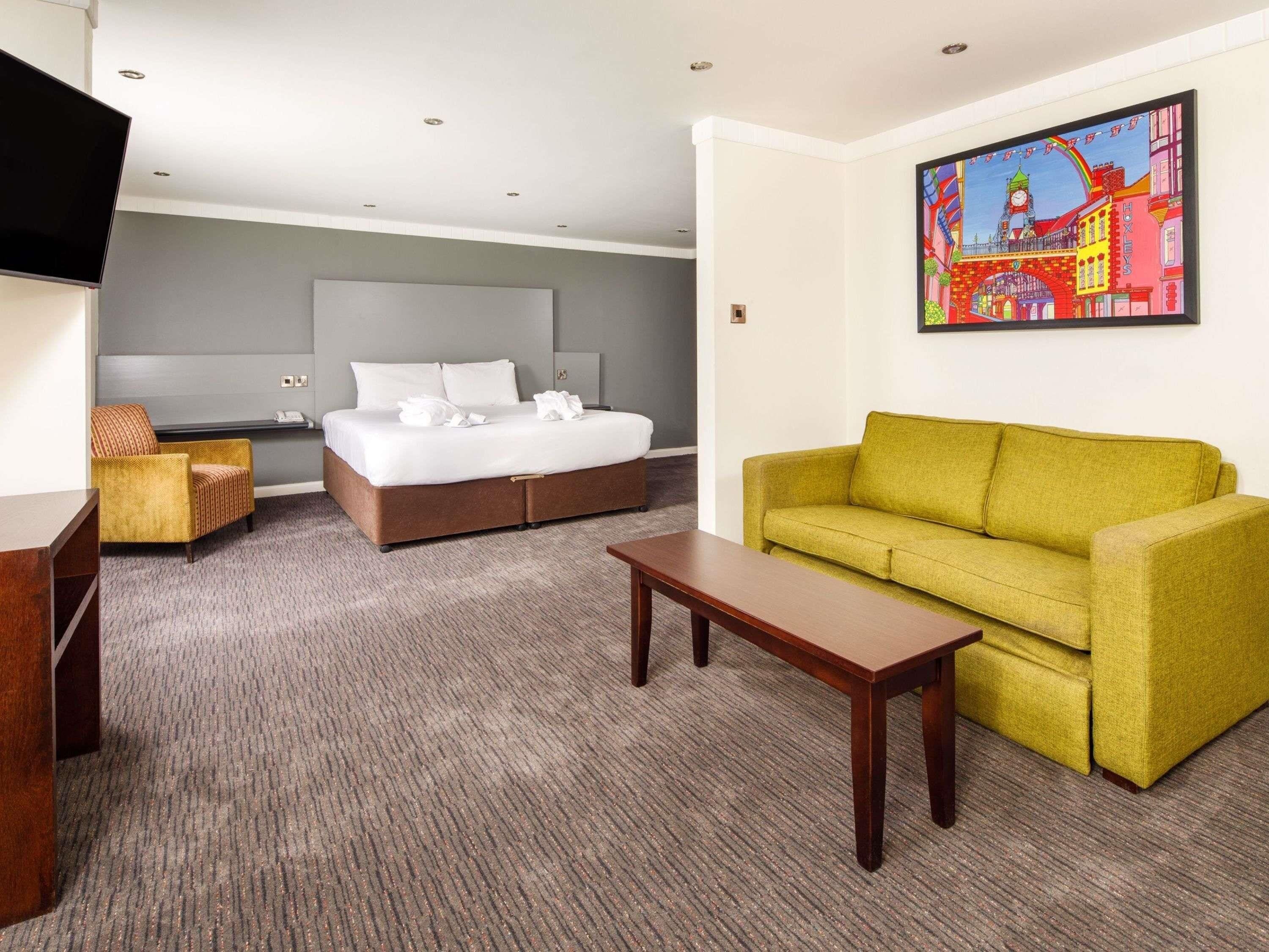 Quarto de hotel com uma cama, um sofá verde, uma mesa de madeira e um quadro na parede.