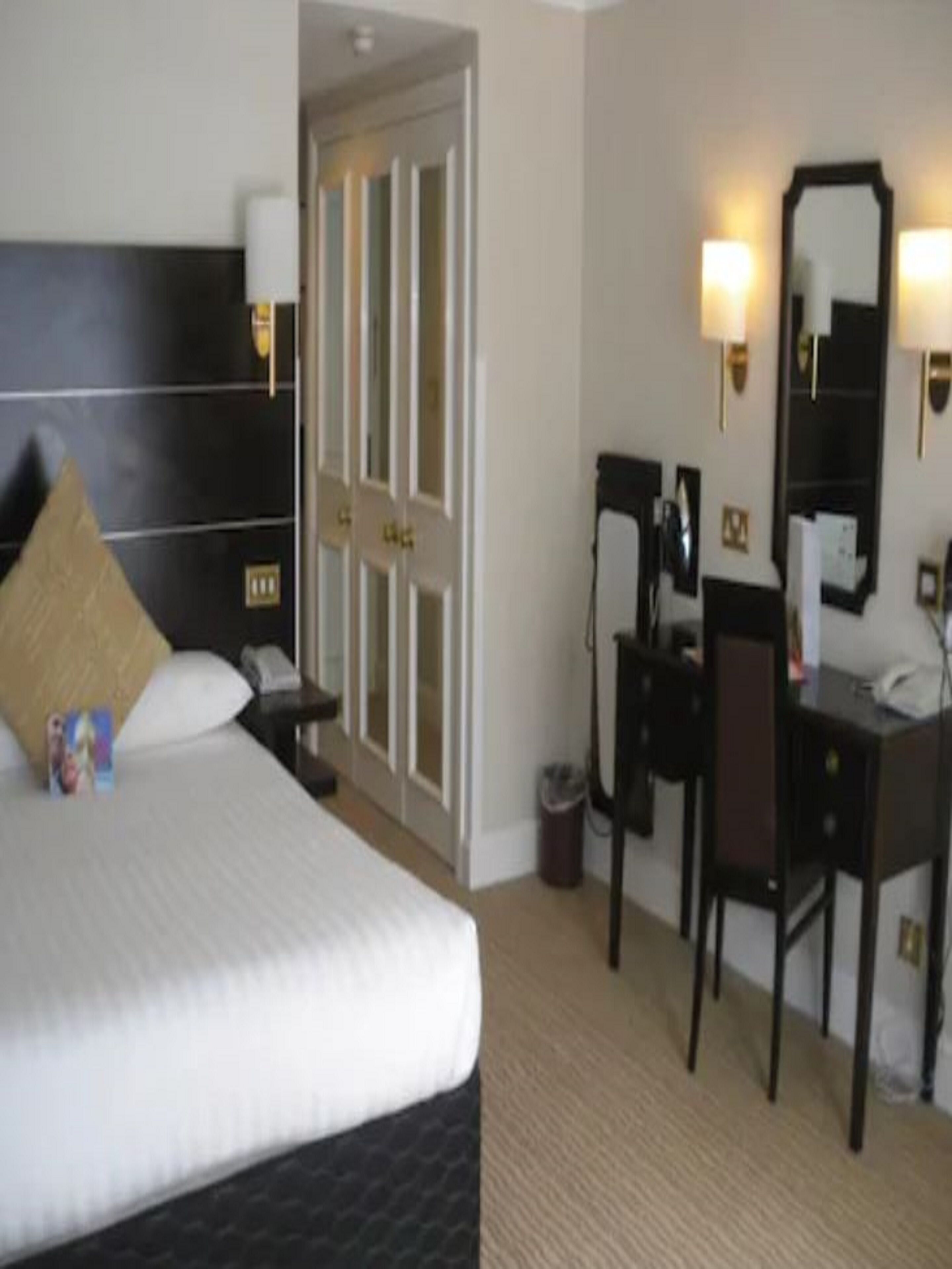 Quarto de hotel com cama, escrivaninha, cadeira, espelho e porta.