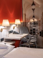Quarto de hotel com cama, escrivaninha com telefone, duas luminárias de parede e uma cadeira.
