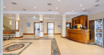 Um saguão de hotel com uma mesa de recepção, um refrigerador repleto de bebidas e uma escadaria para outro andar.