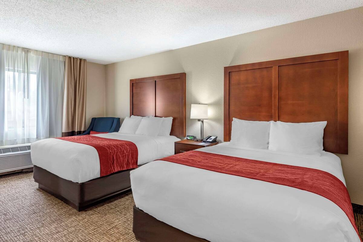 Quarto de hotel com duas camas, cada uma com cabeceira de madeira, roupa de cama branca e um toque de cor vermelha. Há uma mesa de cabeceira com um abajur e um telefone.