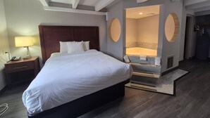 Quarto de hotel com uma cama grande, mesa de cabeceira com abajur, espelho e banheiro com banheira e porta-toalhas.