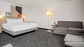 Quarto de hotel com uma cama, um sofá cinzento, uma mesinha e um candeeiro de chão.