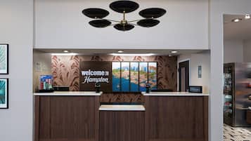 Balcão de recepção com um letreiro 'Welcome to Hampton', um lustre moderno e um mural com vista para a cidade.