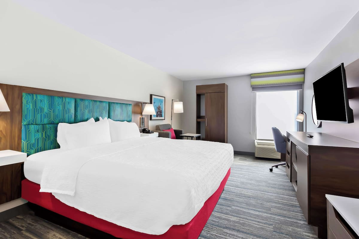 Quarto de hotel com uma cama grande, uma escrivaninha, uma cadeira, uma televisão e uma janela.