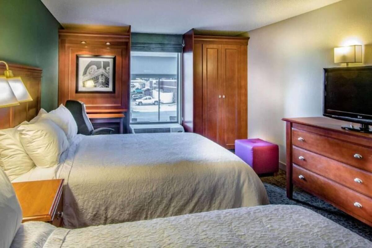 Quarto de hotel com cama, escrivaninha, cadeira, televisão, cômoda e janela com vista para o exterior.