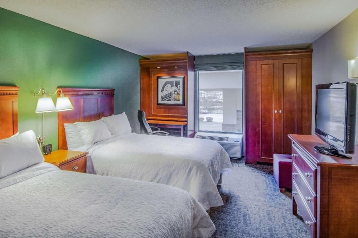 Quarto de hotel com duas camas, uma escrivaninha, televisão e janela.