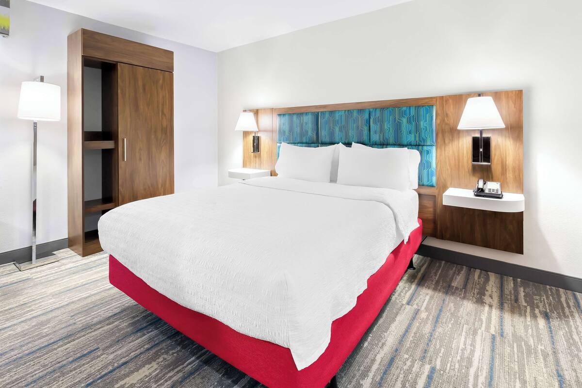 Quarto de hotel com uma cama grande, um guarda-roupa de madeira, duas mesas de cabeceira com luminárias e um tapete estampado.
