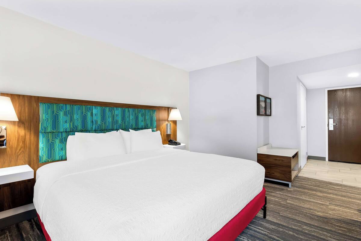 Quarto de hotel com uma cama grande, cabeceira de madeira, duas mesas de cabeceira com luminárias, um pequeno banco de madeira e uma porta.