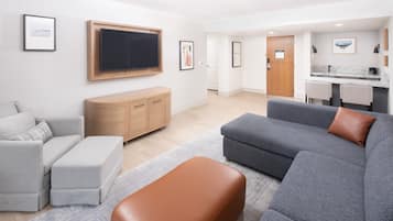 Uma sala de estar moderna com um sofá cinzento, um banquinho marrom e um sofá cinza. Há um armário de madeira, uma TV fixada na parede e uma área de cozinha ao fundo.