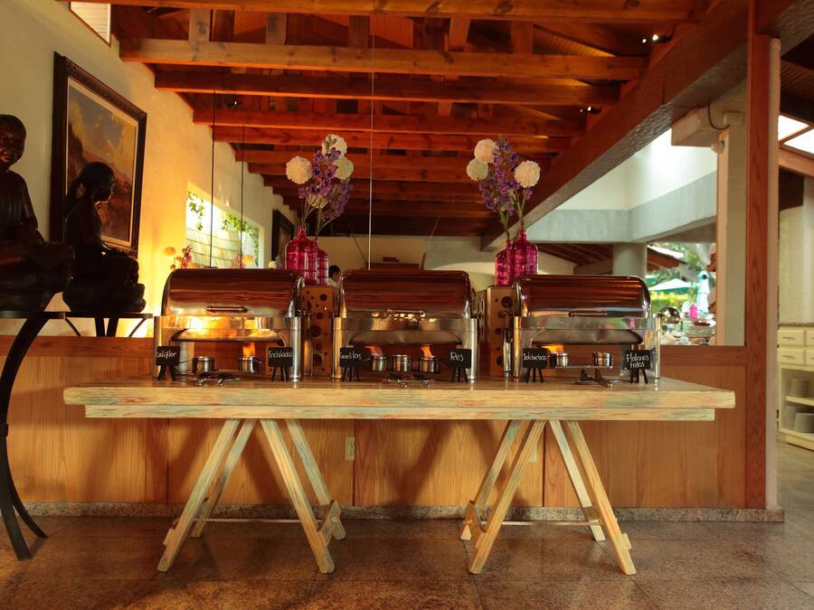 Uma área de bar com balcão comprido, teto rústico e vasos decorativos com flores.