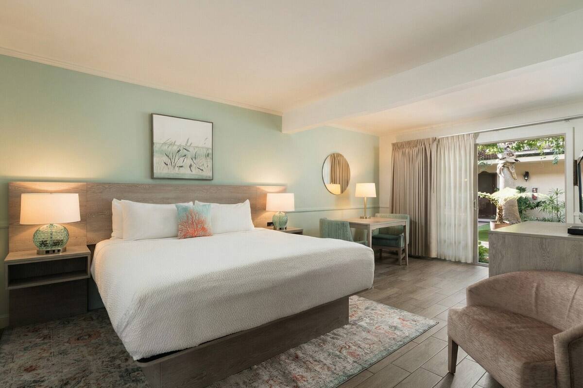 Quarto de hotel com uma cama grande, mesas de cabeceira, escrivaninha, cadeira e vista para o exterior.