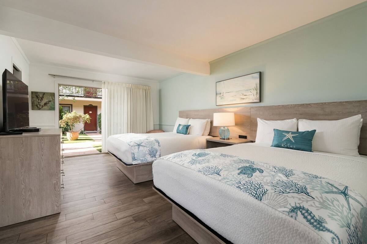 Quarto de hotel com duas camas, televisão, escrivaninha e vista para a área externa.