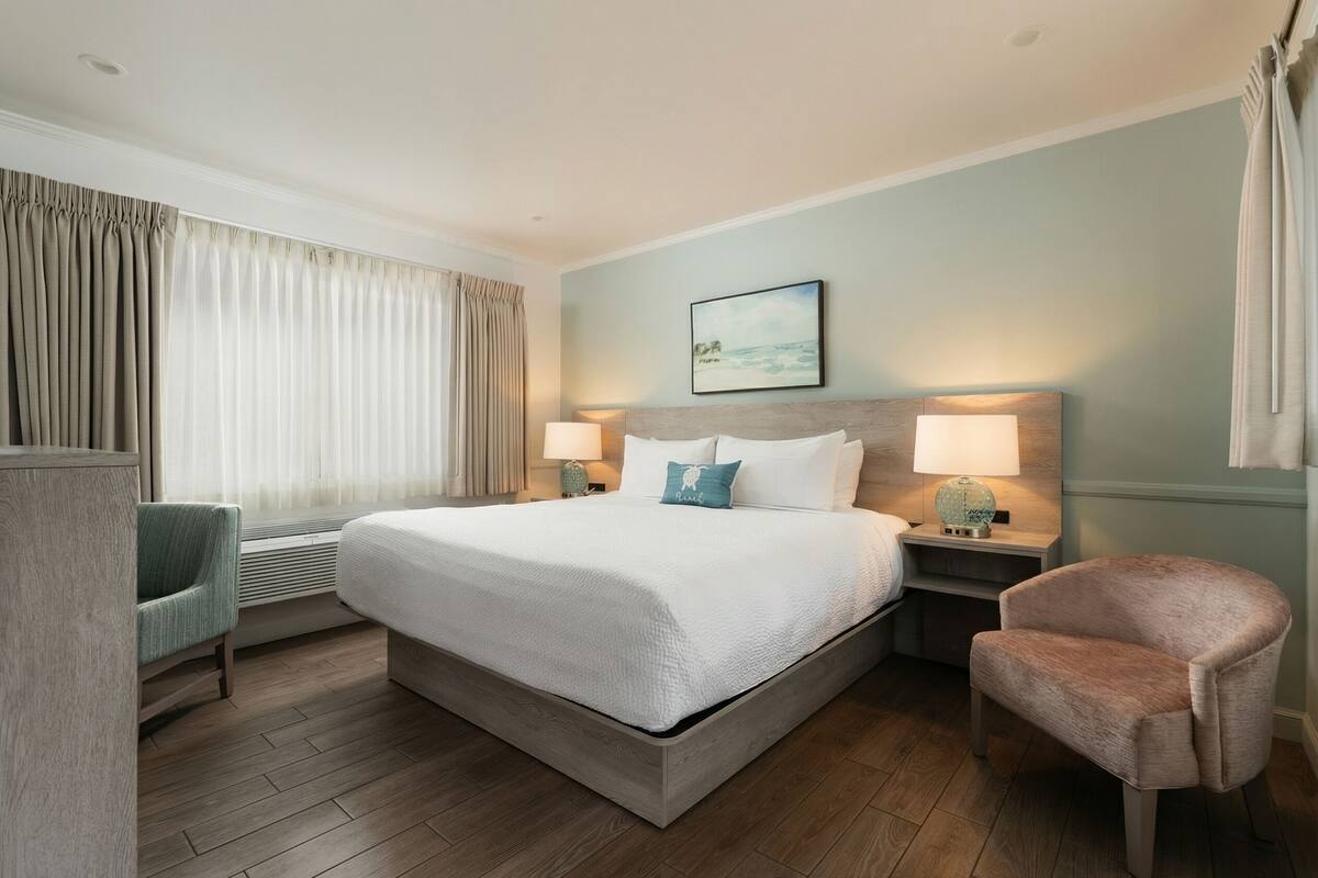 Quarto de hotel com uma cama grande, duas mesas de cabeceira com luminárias, uma cadeira e uma janela com cortinas.