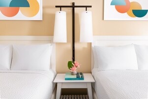 Quarto de hotel com duas camas, uma pequena mesa de cabeceira com um livro e um vaso de flores, e duas luminárias fixadas na parede.
