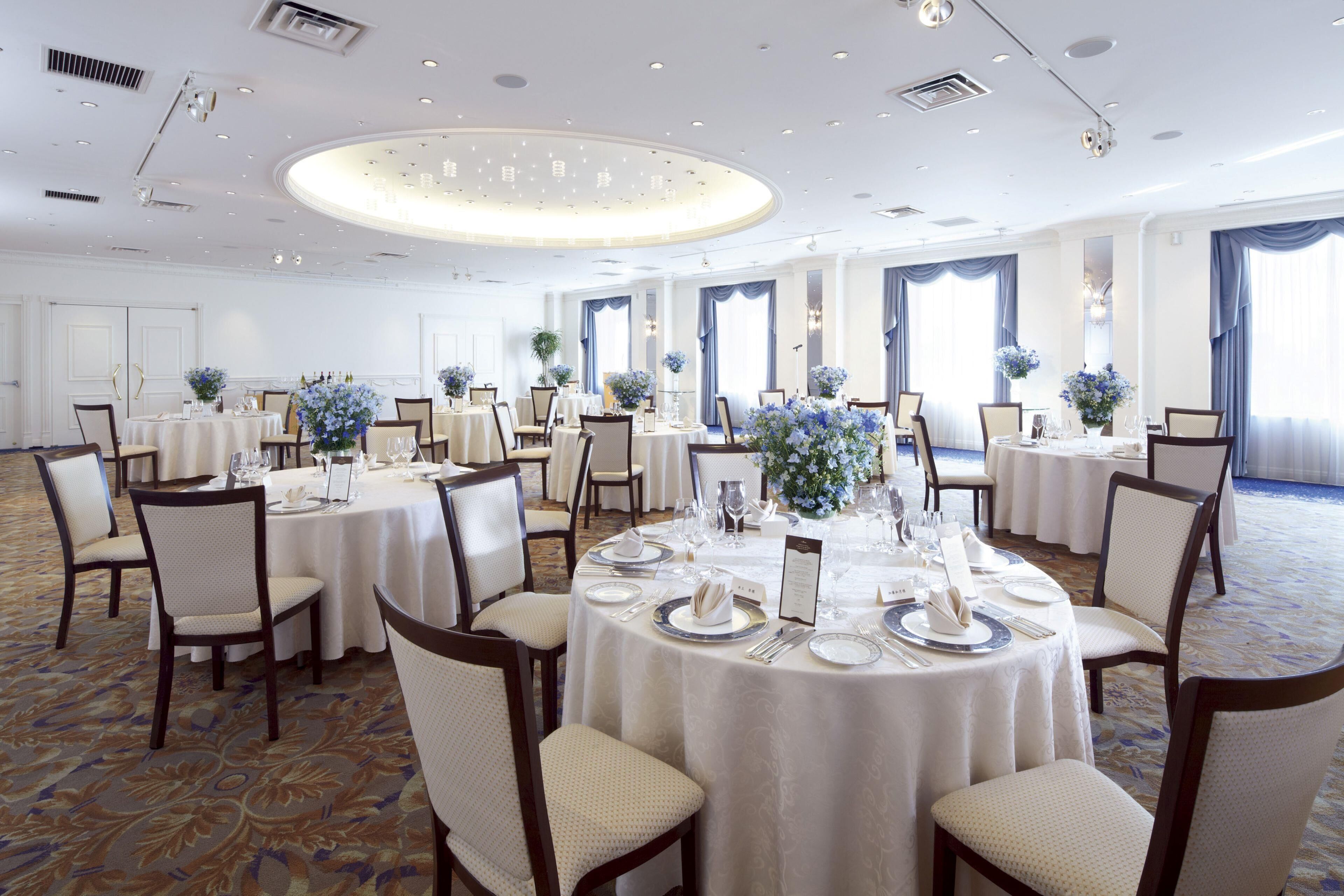 Uma sala de jantar de hotel com mesas redondas, cadeiras e centros de mesa com flores.