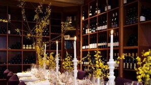 Uma longa mesa de jantar posta com toalhas brancas, velas e flores amarelas, rodeada por cadeiras roxas em um salão com estantes de madeira repletas de garrafas de vinho.