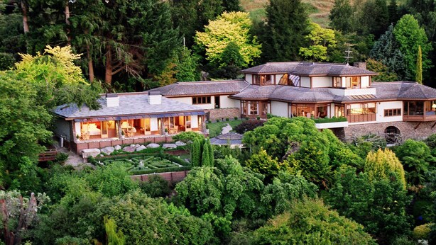 Uma casa grande, de vários andares, rodeada por vegetação densa, com várias janelas e um jardim bem cuidado.