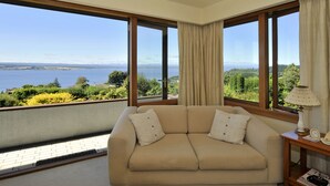 Sala de estar com sofá bege, duas almofadas, mesa lateral com abajur e vista para a água e vegetação através de grandes janelas.