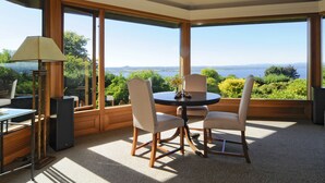 Quarto de canto com uma grande janela que oferece vista para áreas verdes e uma paisagem distante. Há uma mesa redonda com cadeiras, um abajur e um alto-falante.