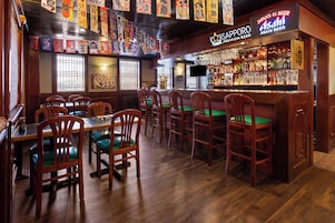 Interior de restaurante com mesas e cadeiras de madeira, área de bar com um anúncio de cerveja Sapporo e várias bandeiras penduradas no teto.