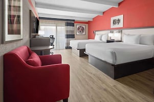 Quarto de hotel com uma parede de destaque vermelha, duas camas, uma poltrona vermelha, uma escrivaninha com abajur e uma televisão.