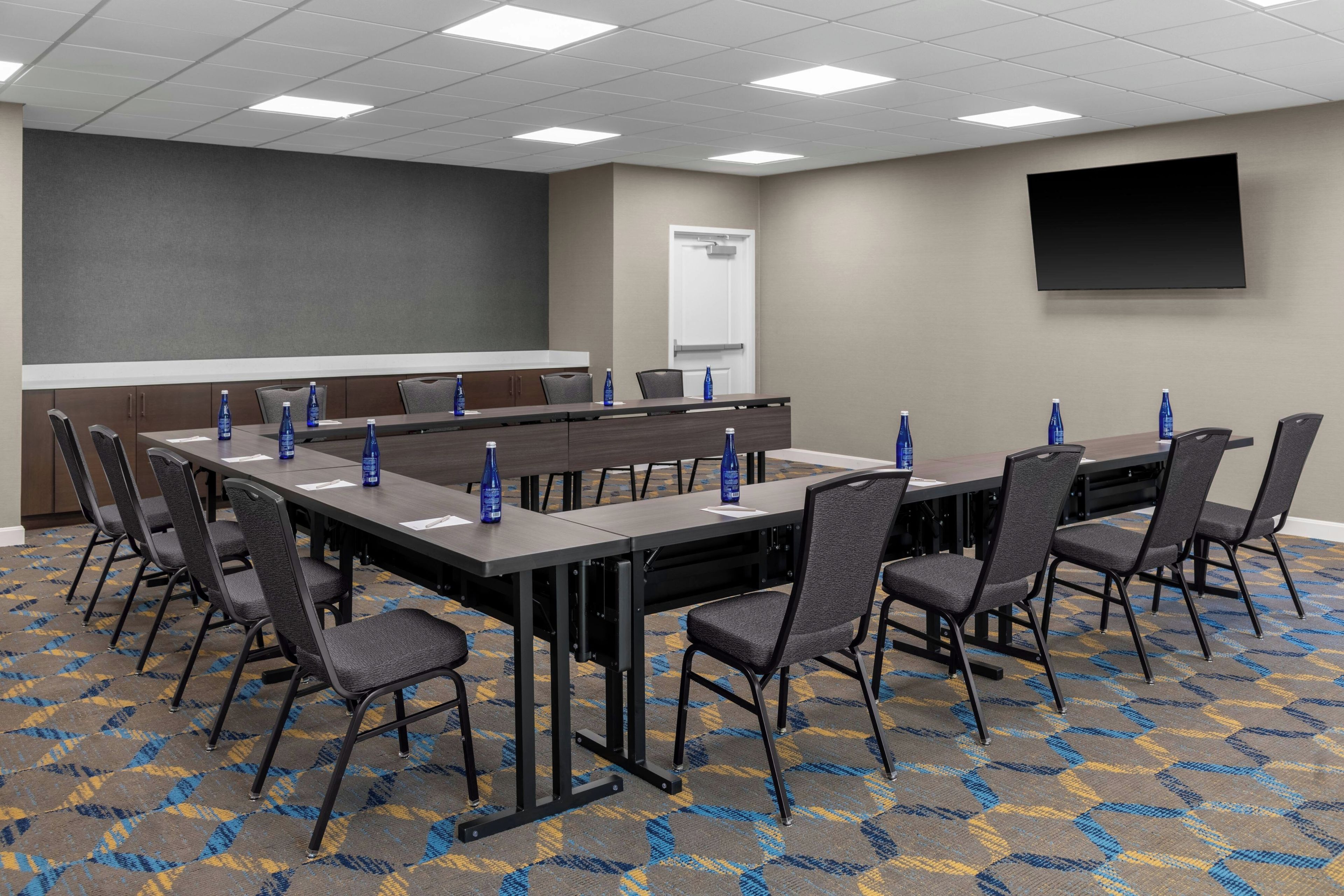 Sala de conferências com uma mesa grande, cadeiras, garrafas de água, tela de projetor e piso coberto de carpete.
