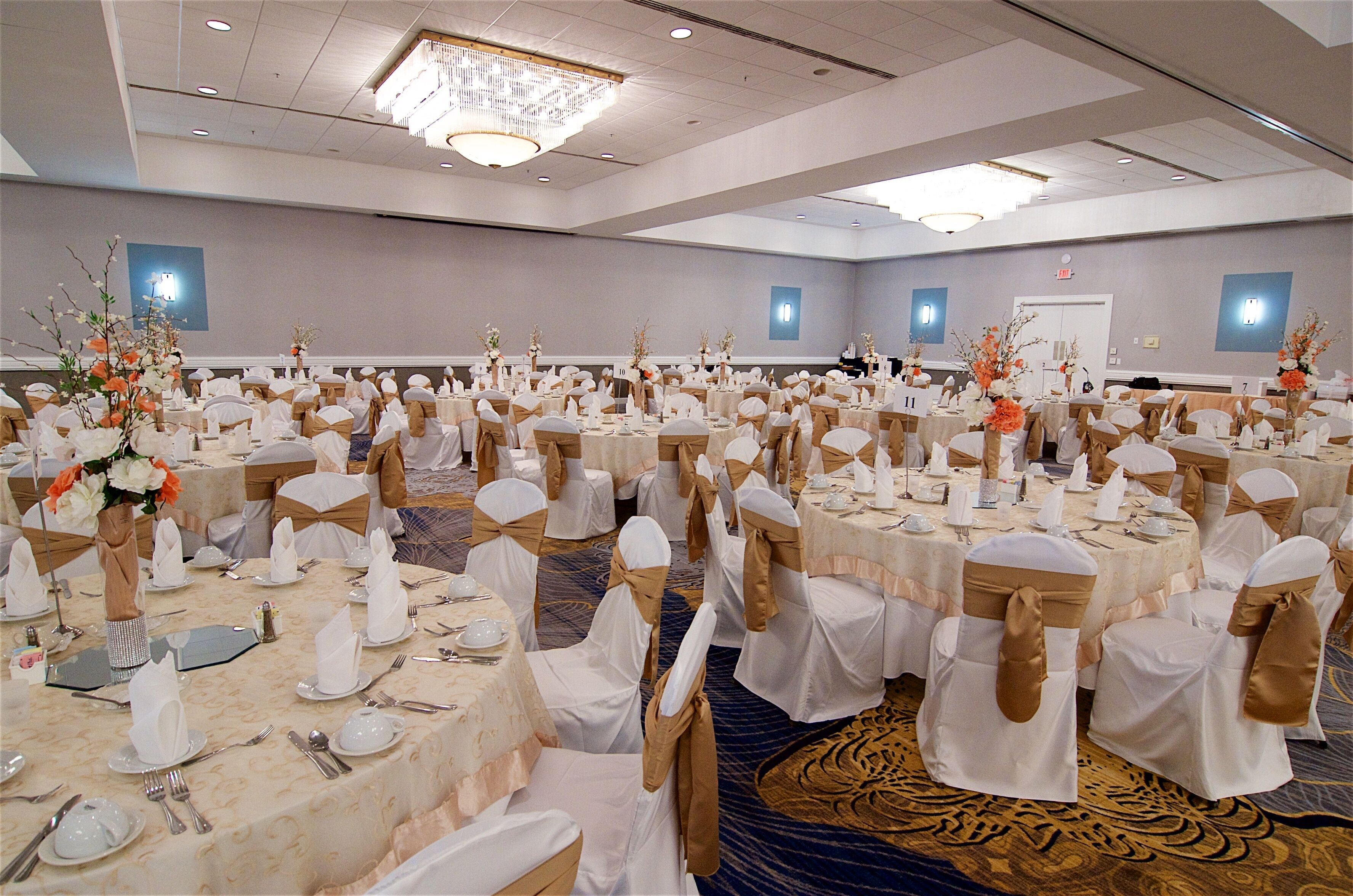 Uma sala de banquetes com mesas redondas dispostas para um evento formal, com toalhas brancas, fitas douradas e arranjos florais.