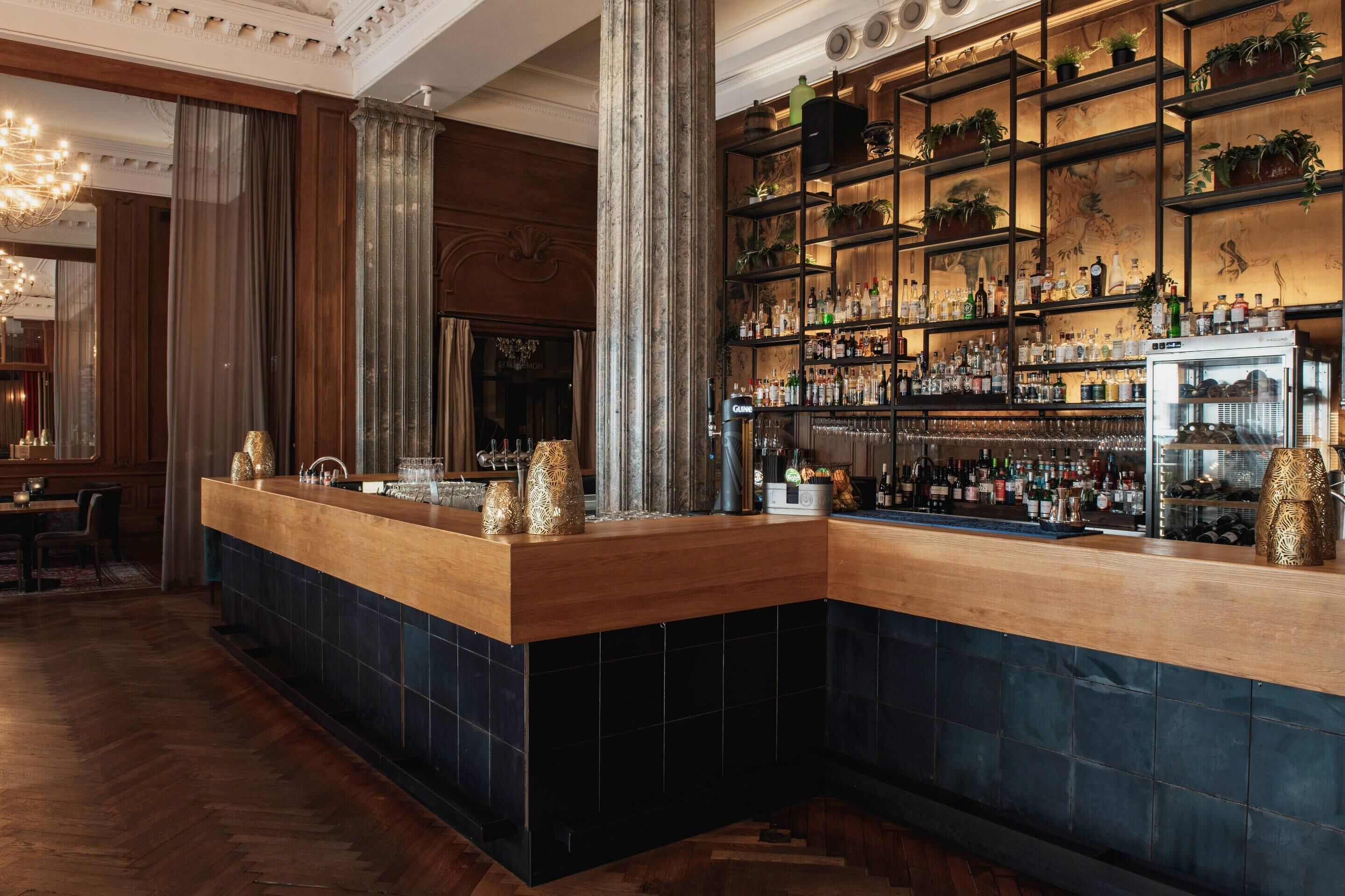 Uma área de bar com balcão de madeira, prateleiras com garrafas e uma parede decorativa com plantas.