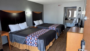 Quarto de hotel com duas camas, piso de madeira, uma escrivaninha, espelho e um pequeno espaço de cozinha.