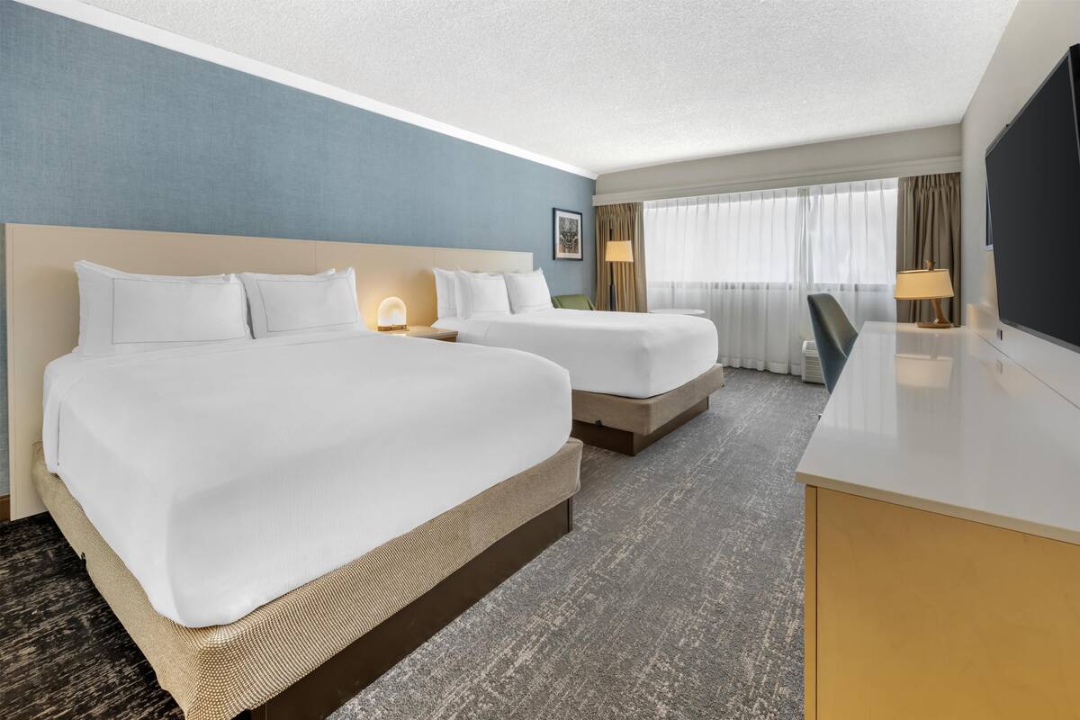 Quarto de hotel com duas camas, uma escrivaninha, uma cadeira, uma televisão e uma janela com cortinas.