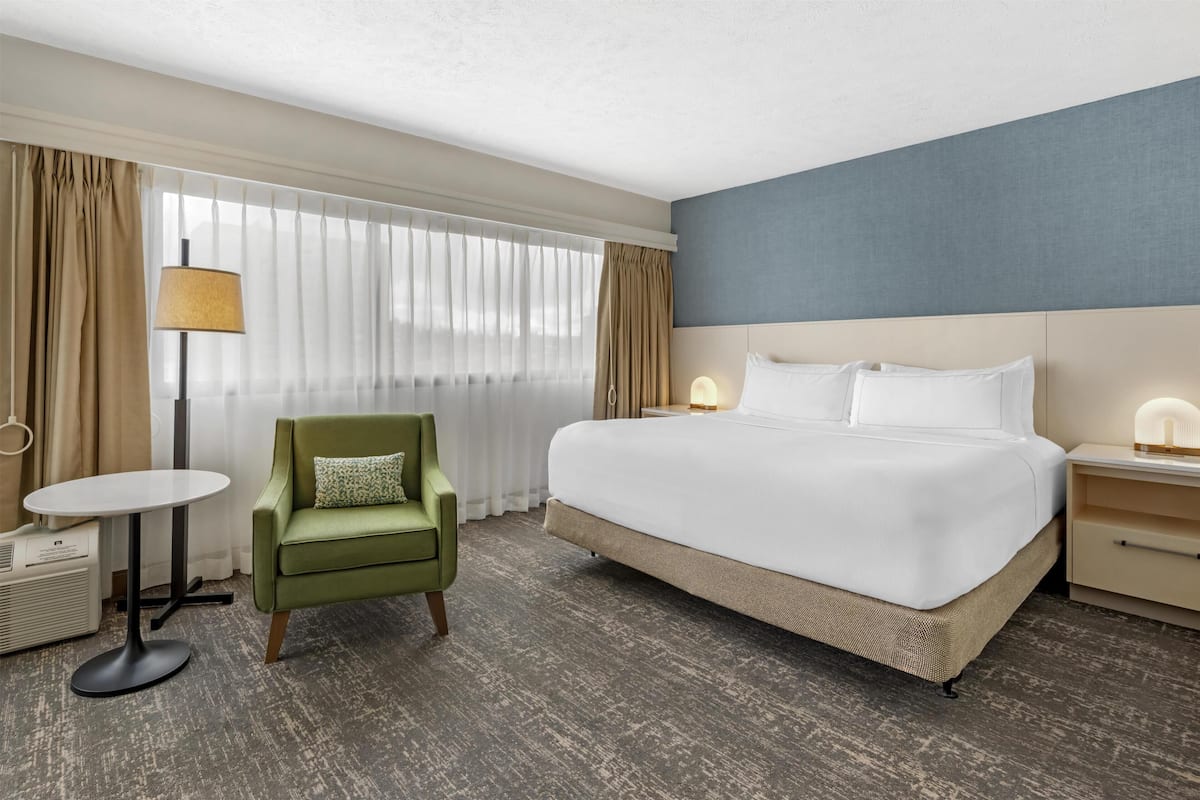 Quarto de hotel com uma cama grande, uma poltrona verde, uma mesa pequena, uma luminária e uma mesa de cabeceira.