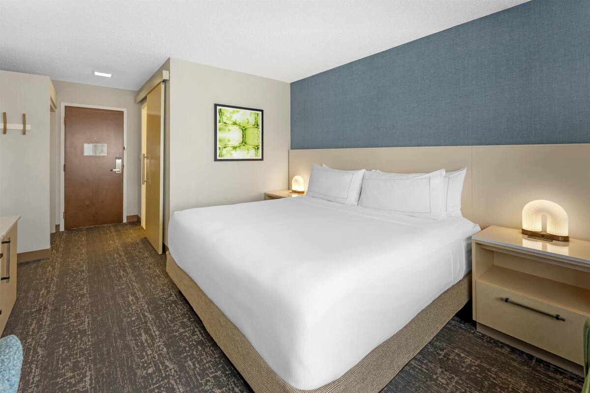 Quarto de hotel com cama grande, mesas de cabeceira, porta e quadro pendurado na parede.