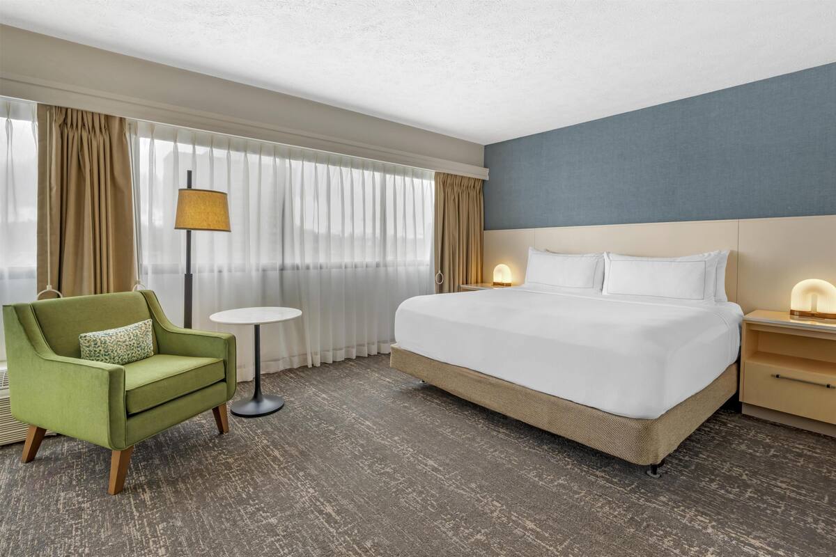 Quarto de hotel com uma cama, uma poltrona verde, uma mesinha redonda e um abajur.