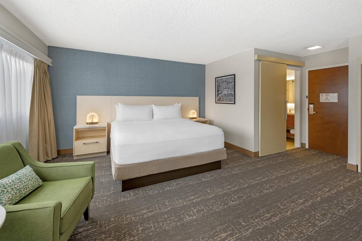 Quarto de hotel com cama, mesas de cabeceira, uma poltrona verde e porta que dá para um banheiro.