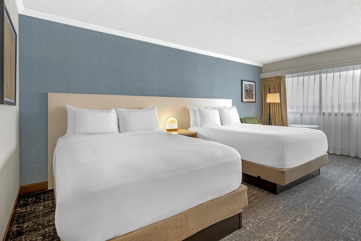 Quarto de hotel com duas camas, uma mesa de cabeceira com abajur, uma janela com cortinas e um quadro na parede.