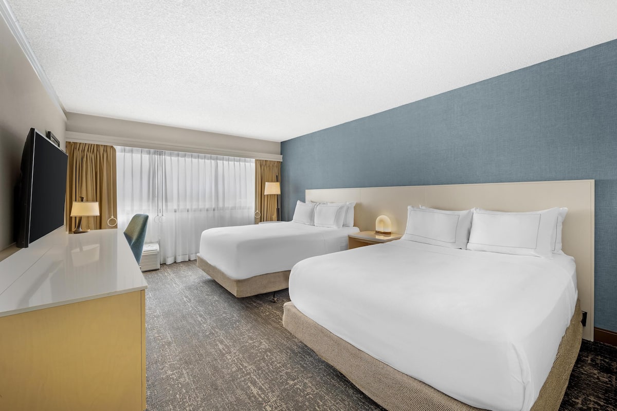 Quarto de hotel com duas camas, uma escrivaninha, uma televisão e uma janela com cortinas.
