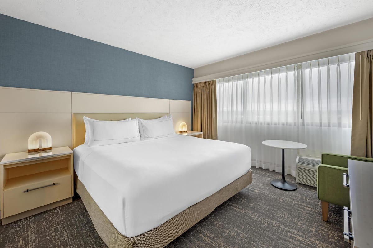 Quarto de hotel com uma cama grande, mesas de cabeceira, uma pequena mesa redonda e uma cadeira.