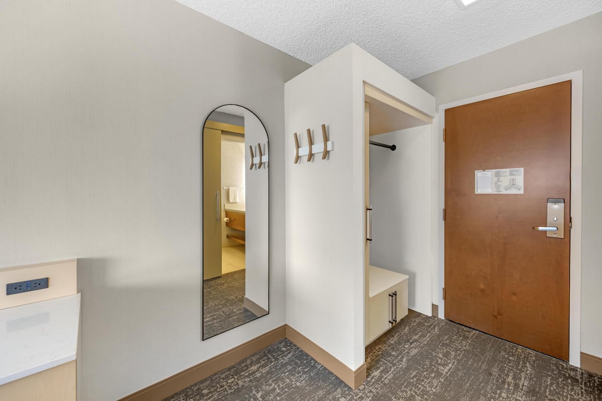 Quarto de hotel com porta de madeira, espelho, pequeno banco e luminária fixada na parede.