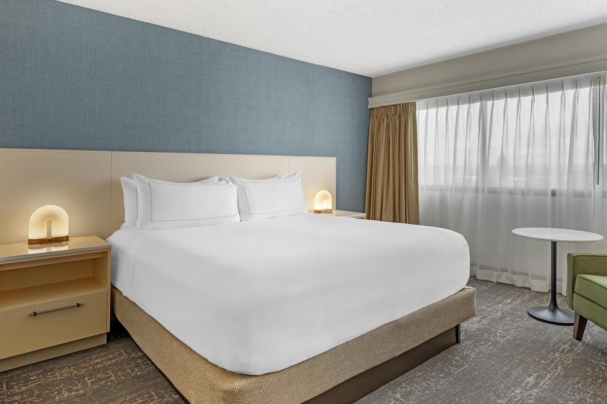 Quarto de hotel com uma cama grande, mesas de cabeceira com abajures, uma pequena mesa redonda e vista para o exterior através de cortinas finas.