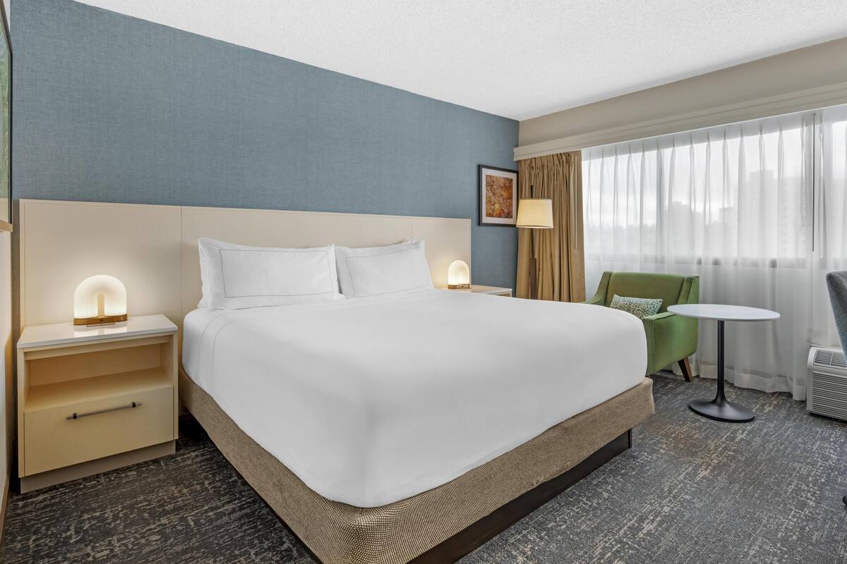Quarto de hotel com uma cama grande, mesas de cabeceira com luminárias, uma cadeira, uma mesinha e uma janela com cortinas.