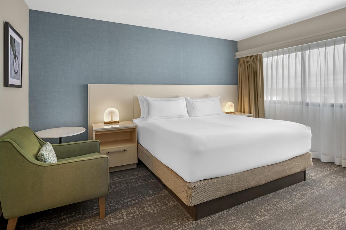 Quarto de hotel com uma cama, uma poltrona verde, uma mesinha de cabeceira e uma luminária na parede.