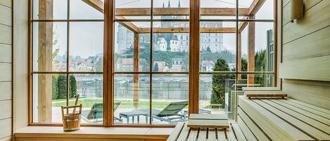 Quarto com amplas janelas, vigas de madeira e vista para edifícios e vegetação.