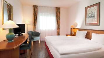 Quarto de hotel com cama, escrivaninha com luminária, cadeira, televisão e quadro na parede.