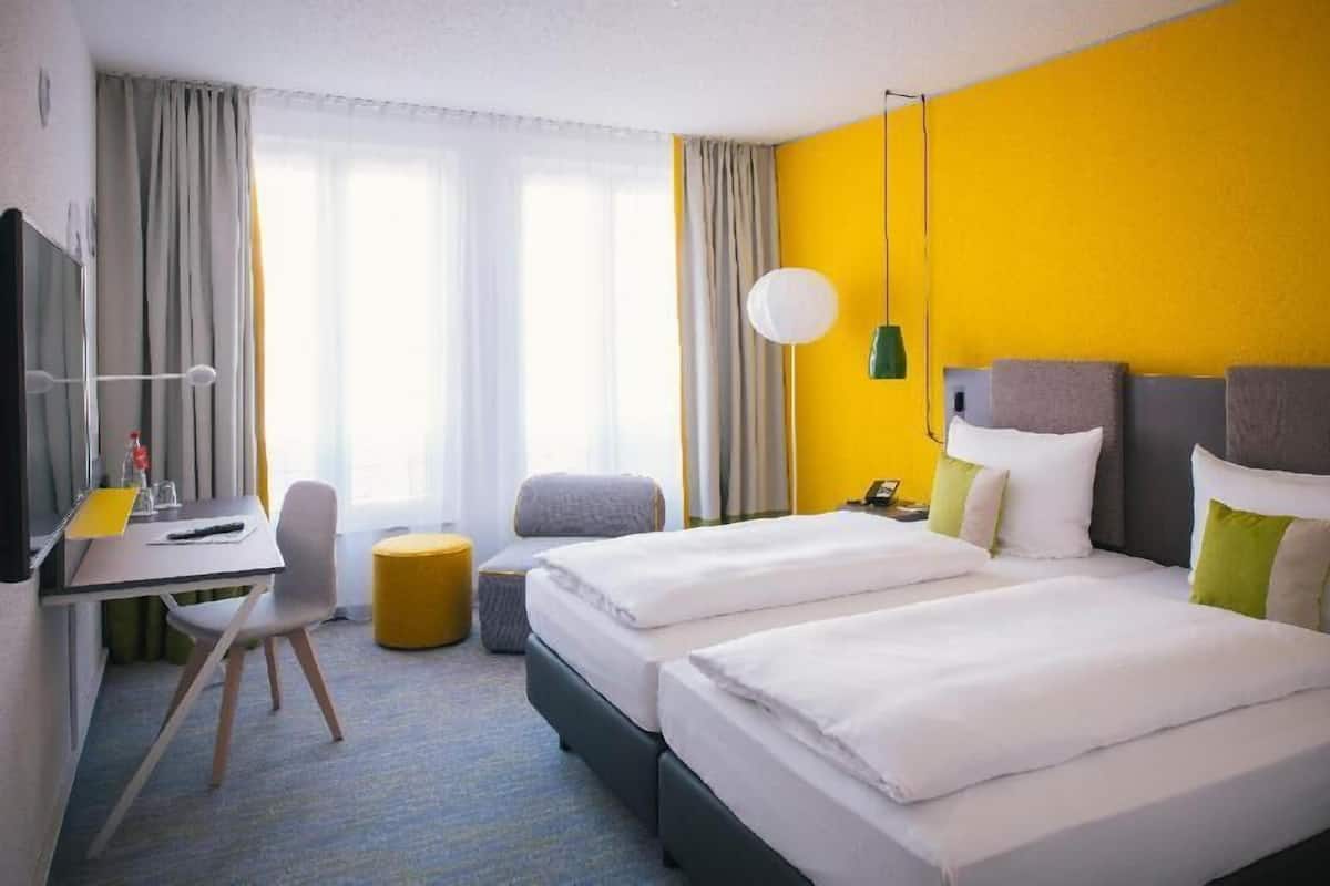 Quarto de hotel com duas camas, uma escrivaninha, uma cadeira, uma luminária e uma janela com cortinas.