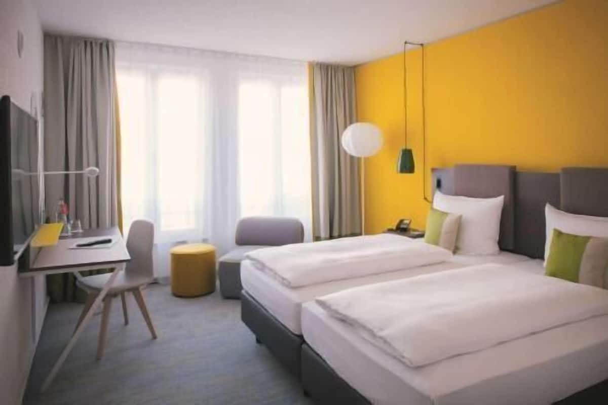 Quarto de hotel com duas camas, uma escrivaninha, uma cadeira, um abajur e uma janela com cortinas.